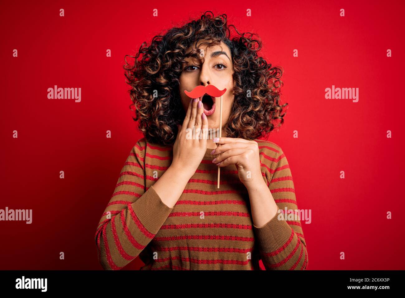 Young beautiful curly arab woman holding funny mustache over isolated ...