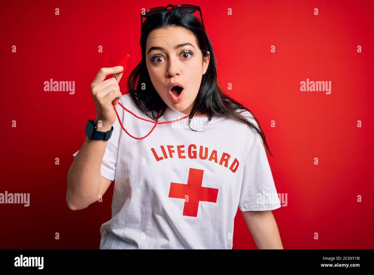 Young lifeguard woman wearing guard equipement holding whistle over red ...