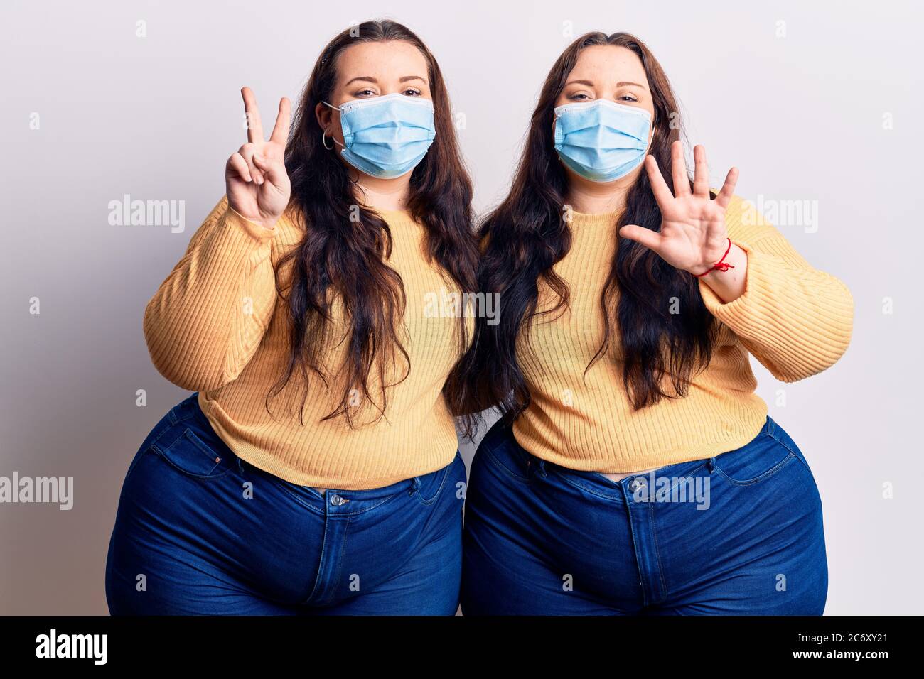 Young plus size twins wearing medical mask showing and pointing up with ...