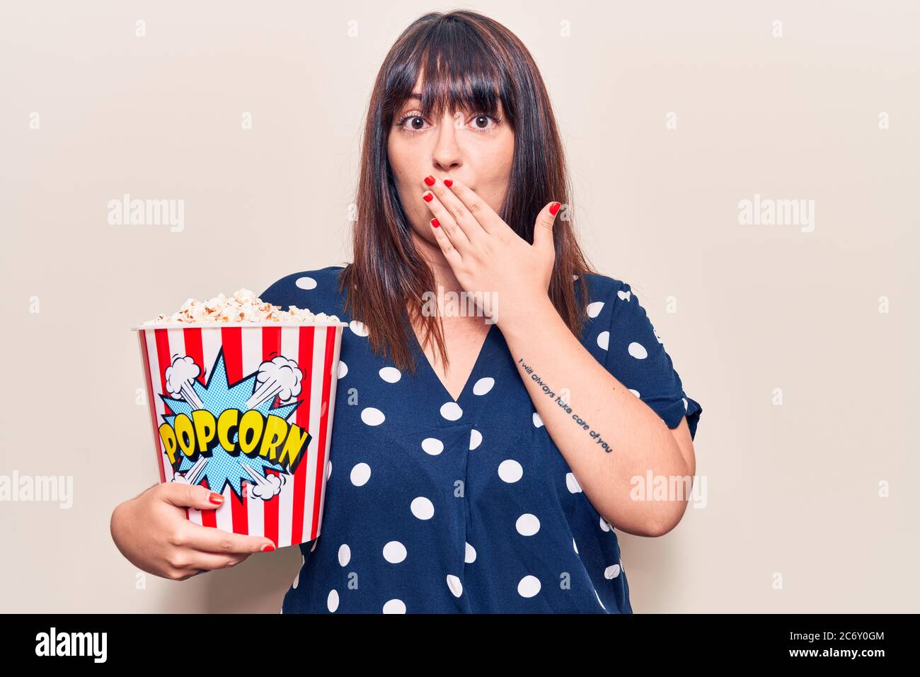 Young plus size woman holding popcorn covering mouth with hand, shocked ...