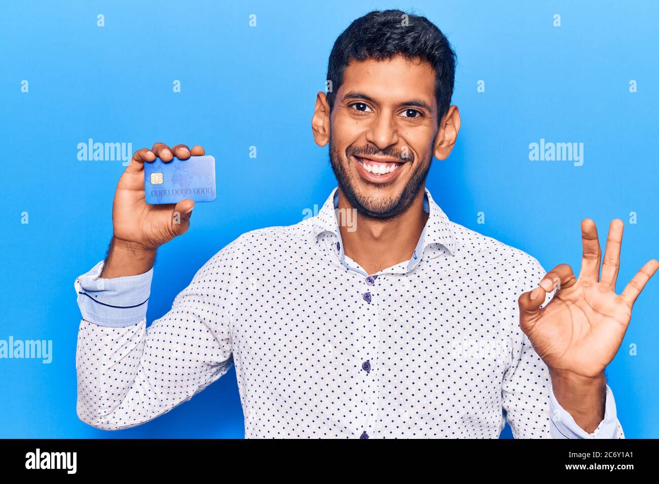 Young latin man holding credit card doing ok sign with fingers, smiling ...