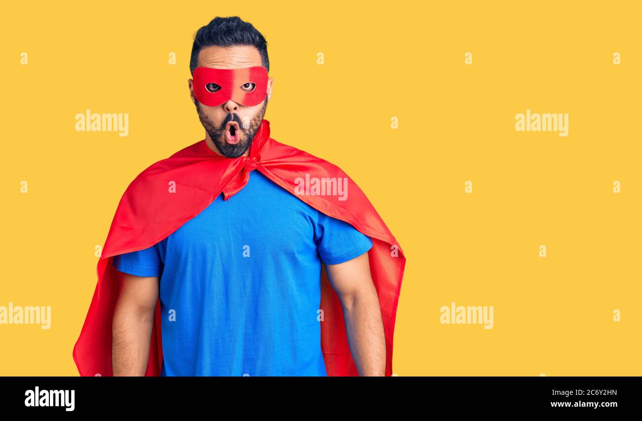 Young hispanic man wearing super hero costume scared and amazed with ...