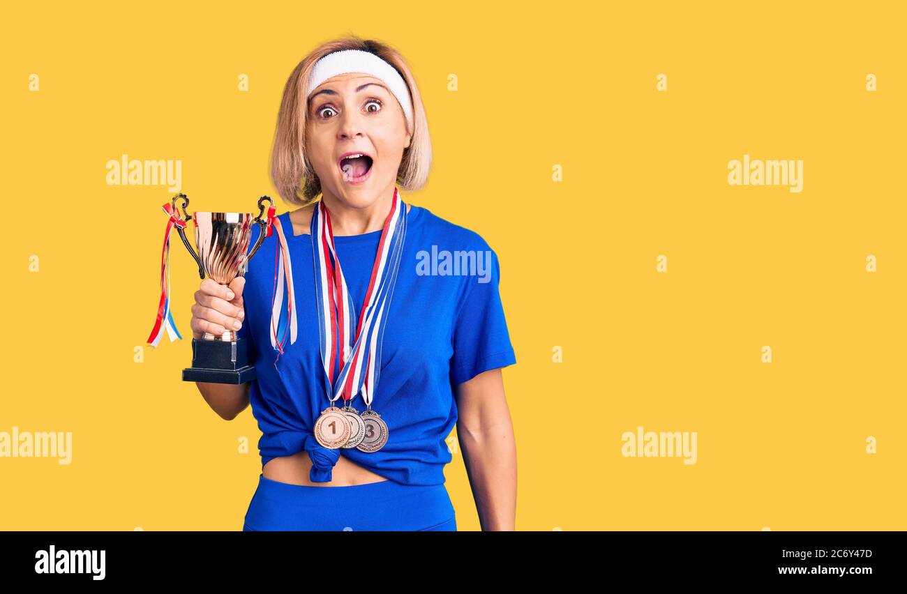Young blonde woman holding champion trophy wearing medals scared and ...