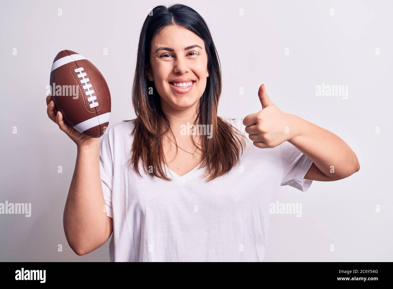 Young beautiful brunette woman playing rubgy holding football ball over ...