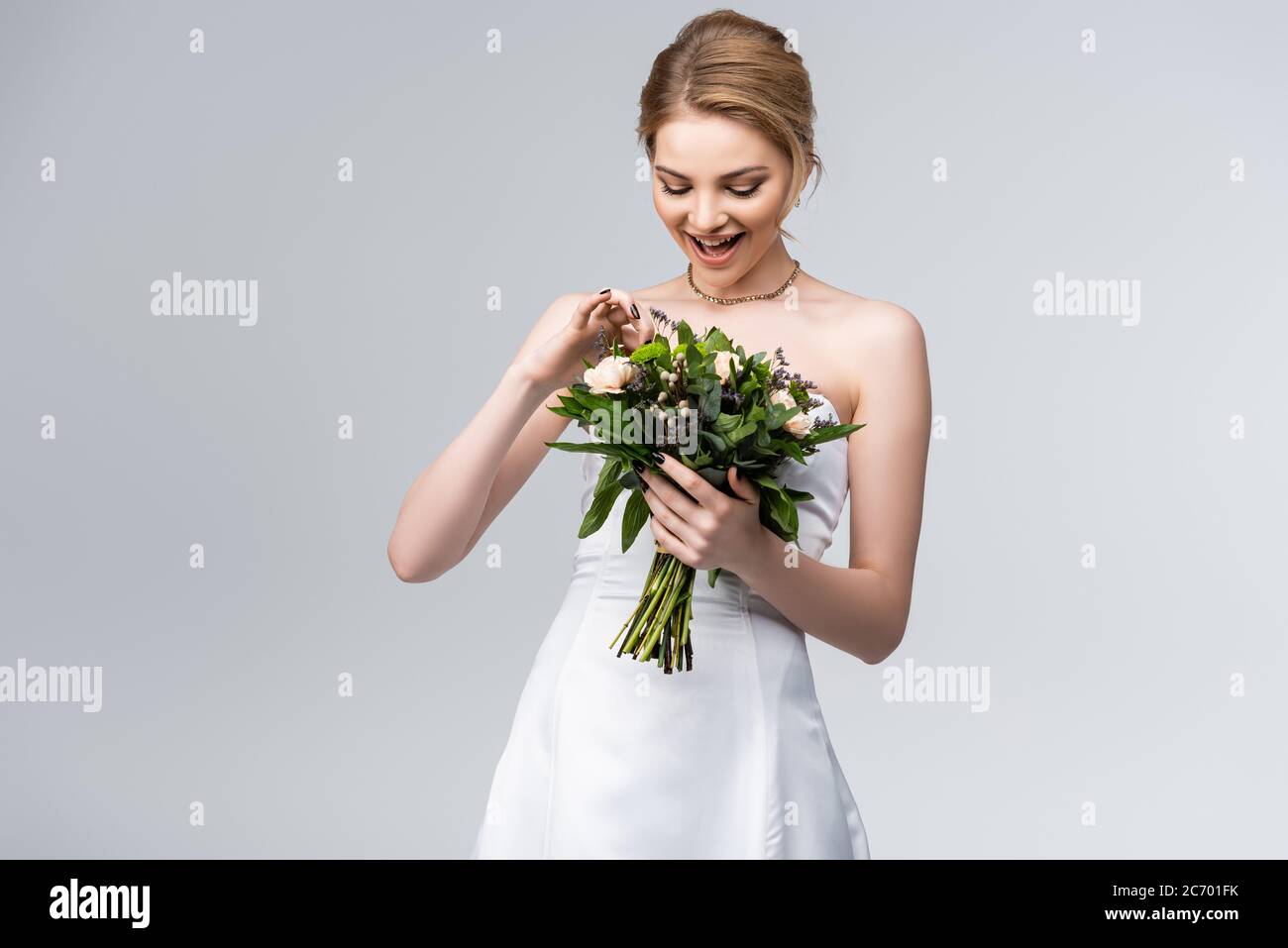 young and excited bride in white dress touching bouquet of flowers ...