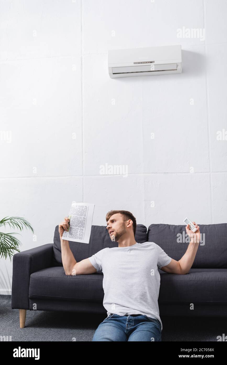 sad young man suffering from heat while using newspaper as hand fan at ...