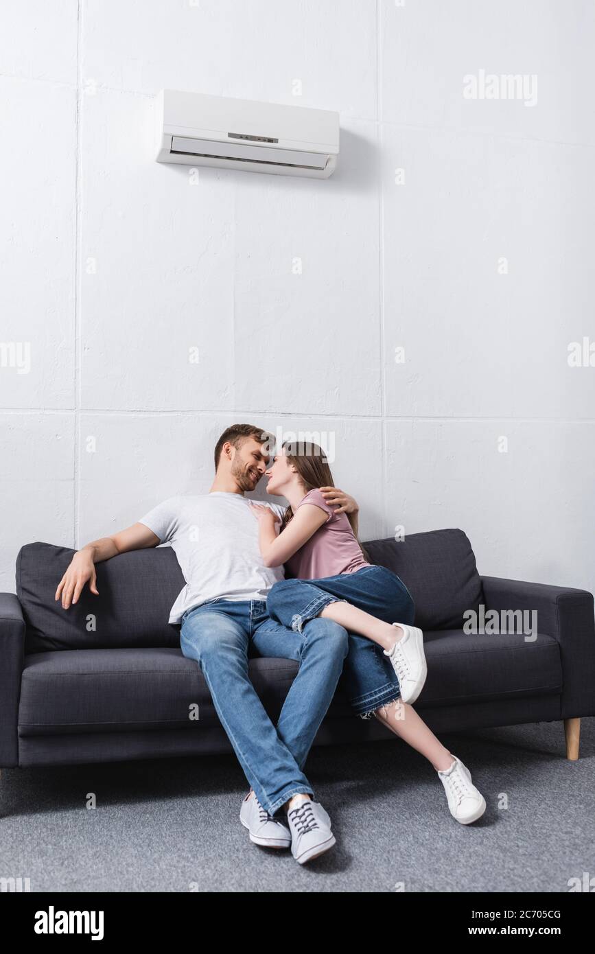 smiling girlfriend and boyfriend hugging and looking at each other at home with air conditioner ...