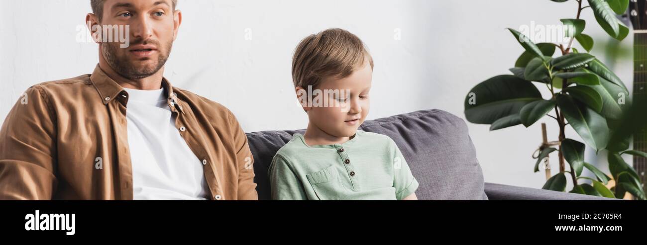 panoramic crop of thoughtful father and son sitting on sofa near potted ...