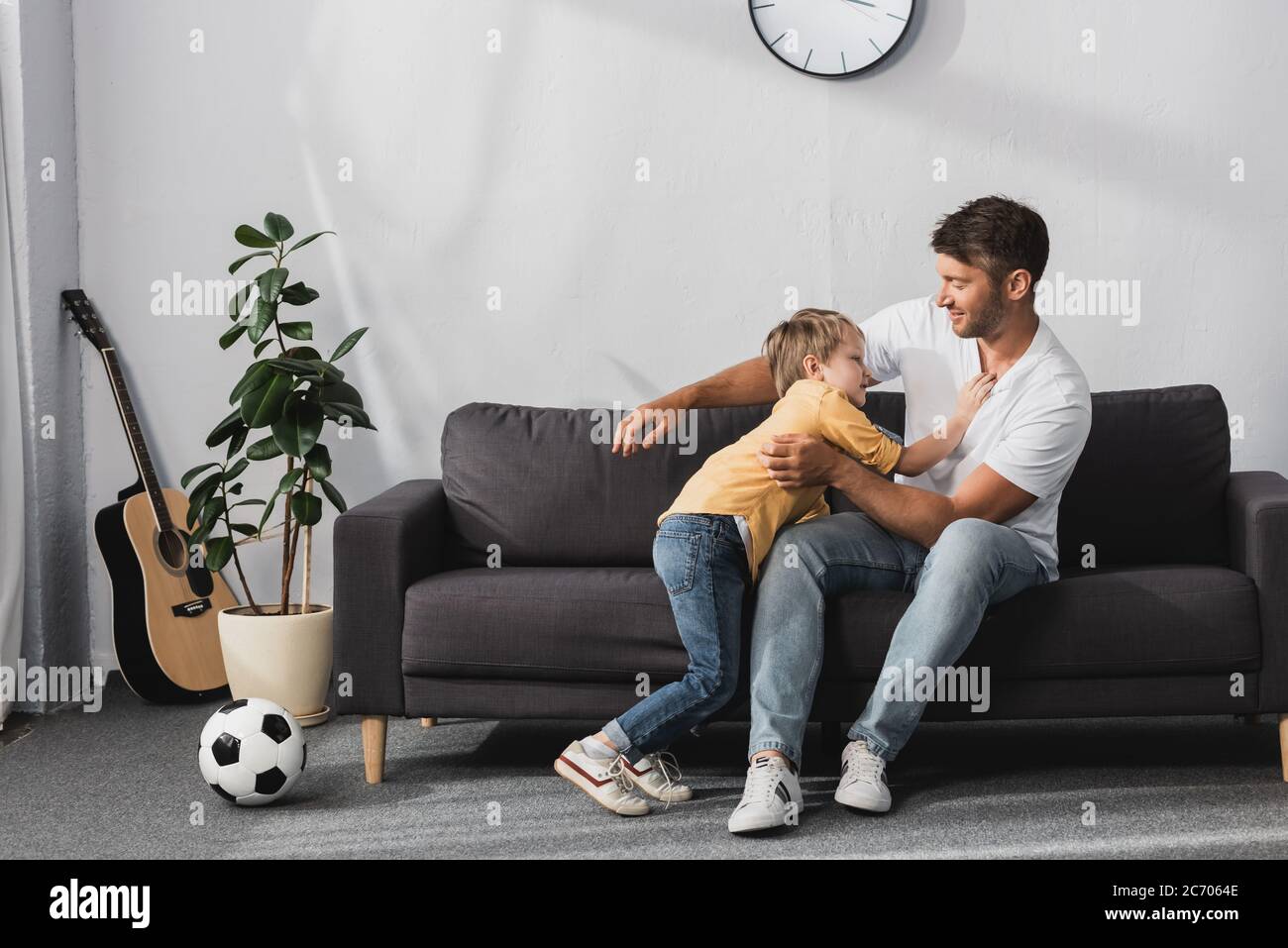 father and adorable son jokingly fighting on sofa near soccer ball and ...