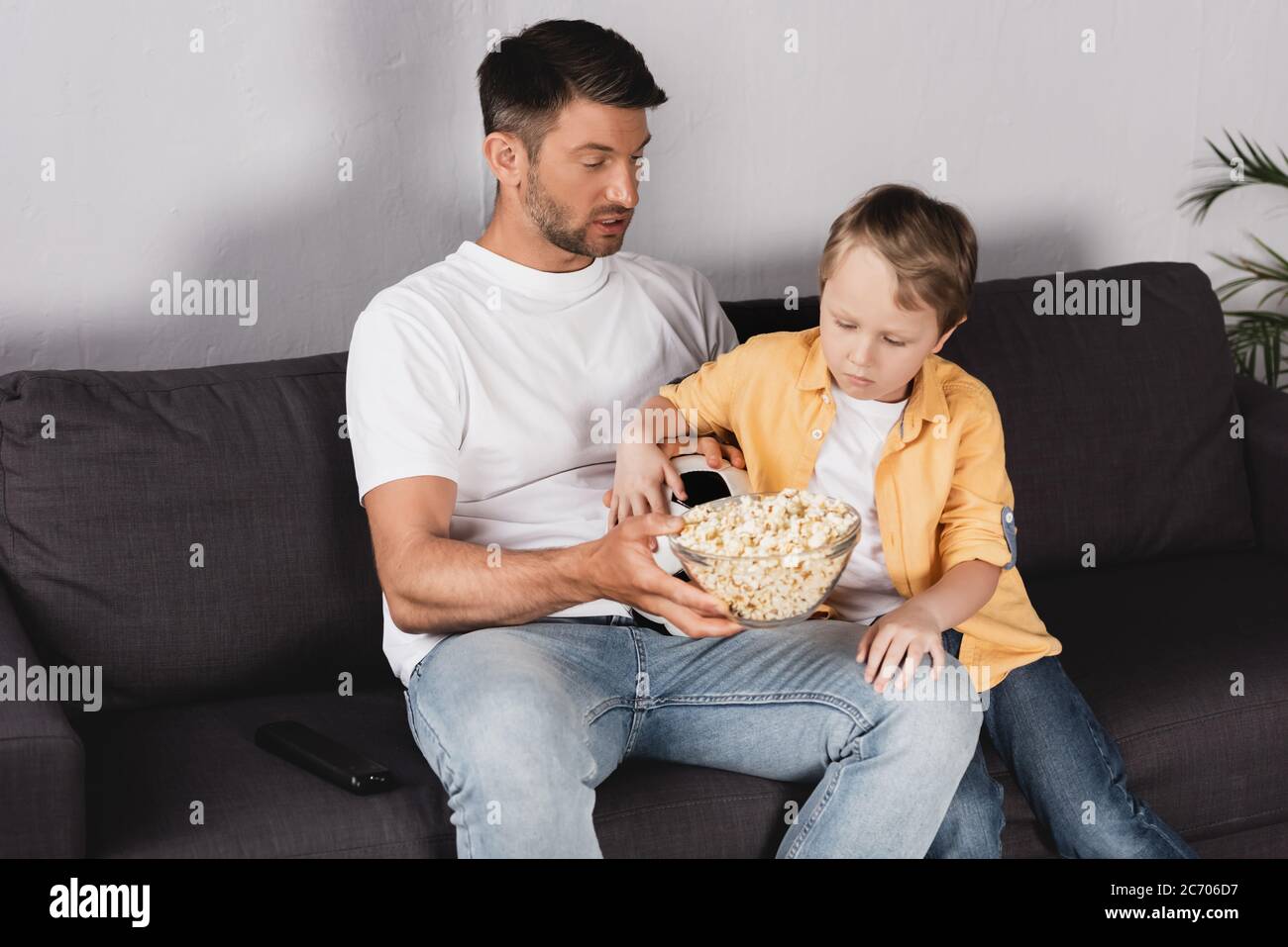 father and son eating popcorn while sitting on sofa and watching tv ...