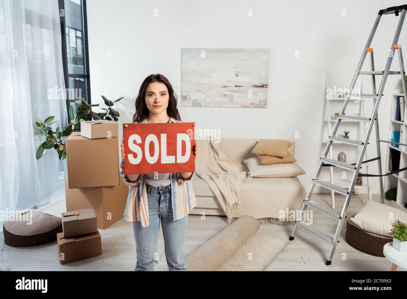 Attractive girl holding nameplate with sold lettering near carton boxes ...