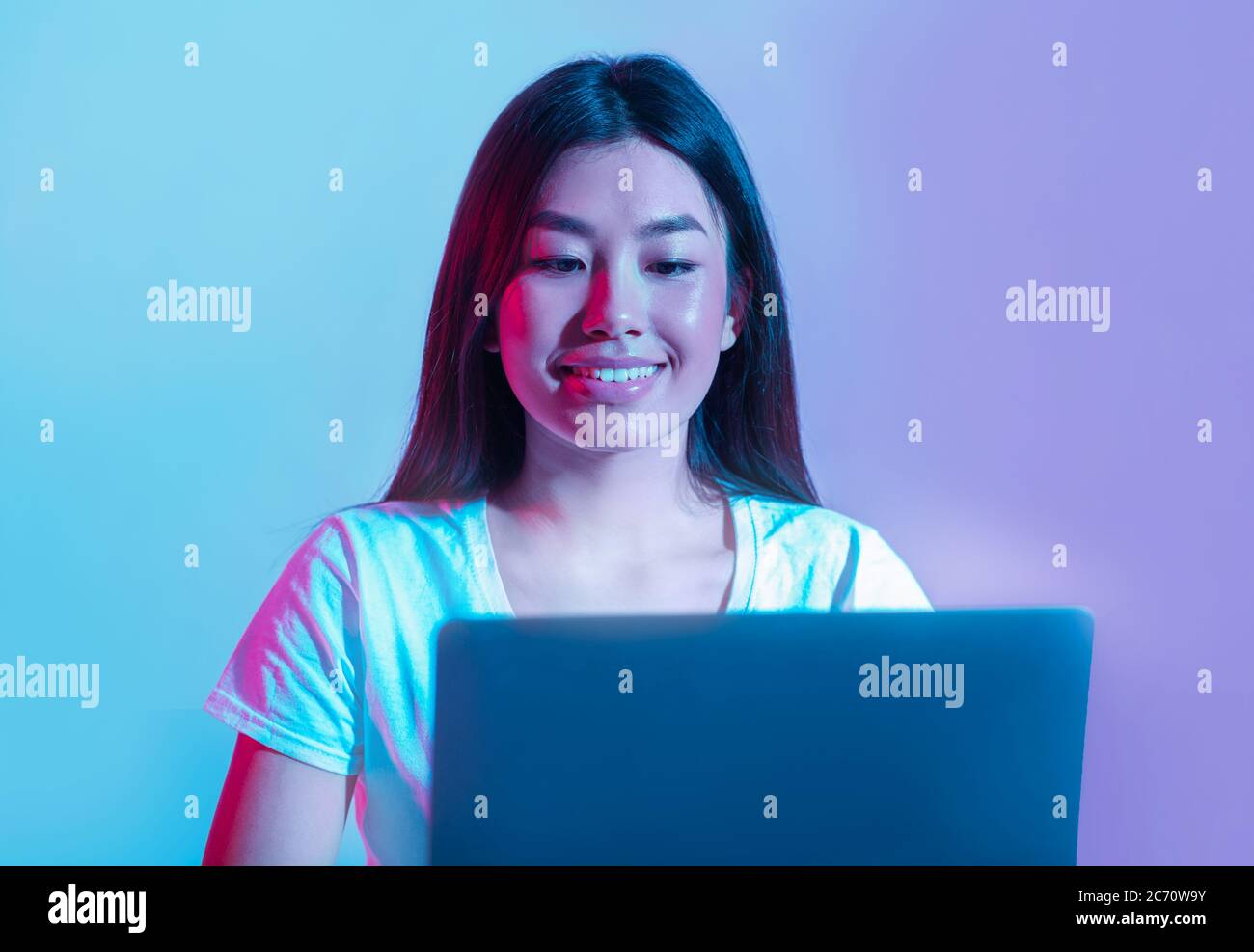 Work or play at computer. Smiling asian girl looking at laptop in neon ...