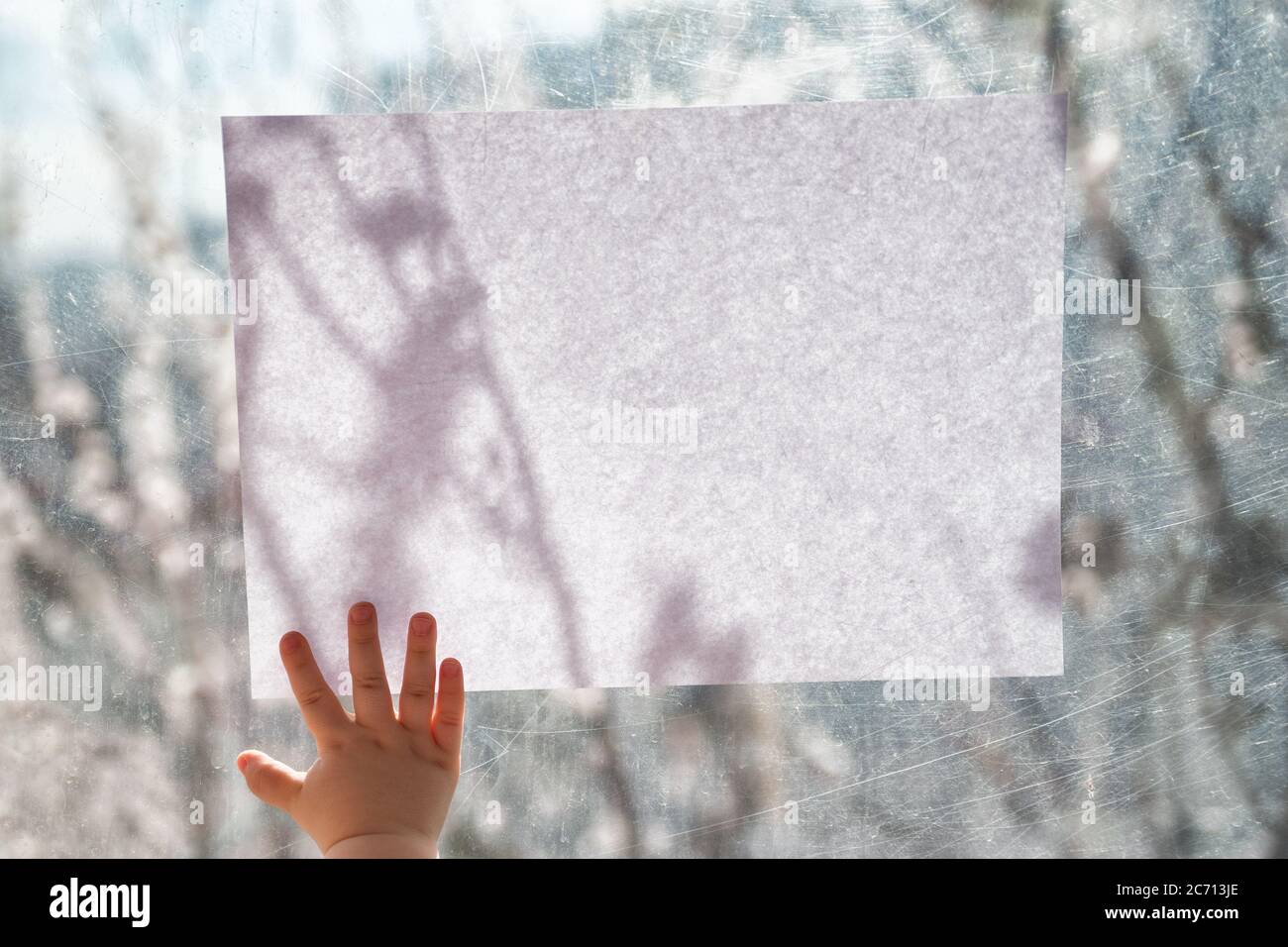 Children's hand holds white sheet of paper on window glass. Place for ...