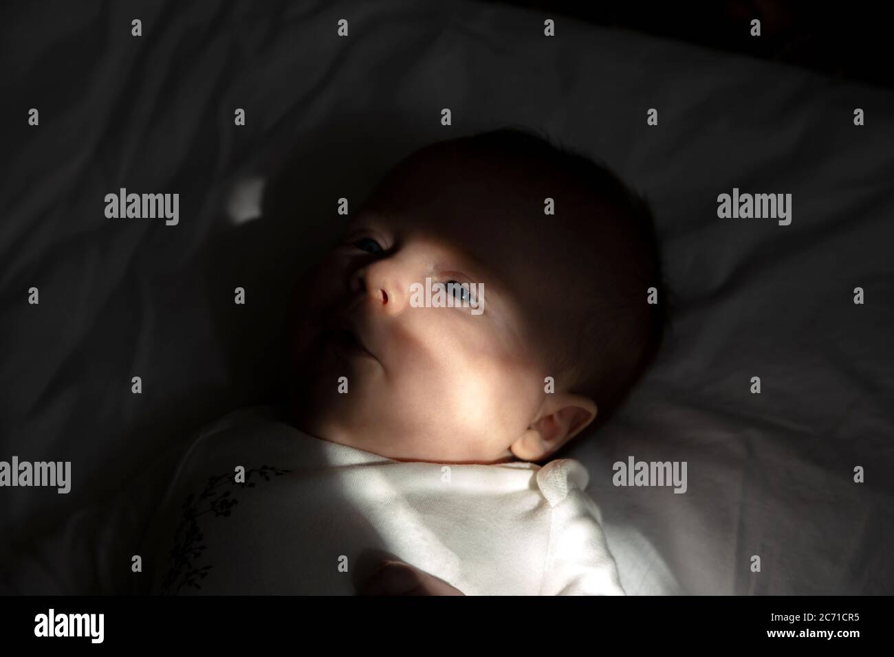 A close-up portrait of a baby girl lying on a bed in the sun. A three ...