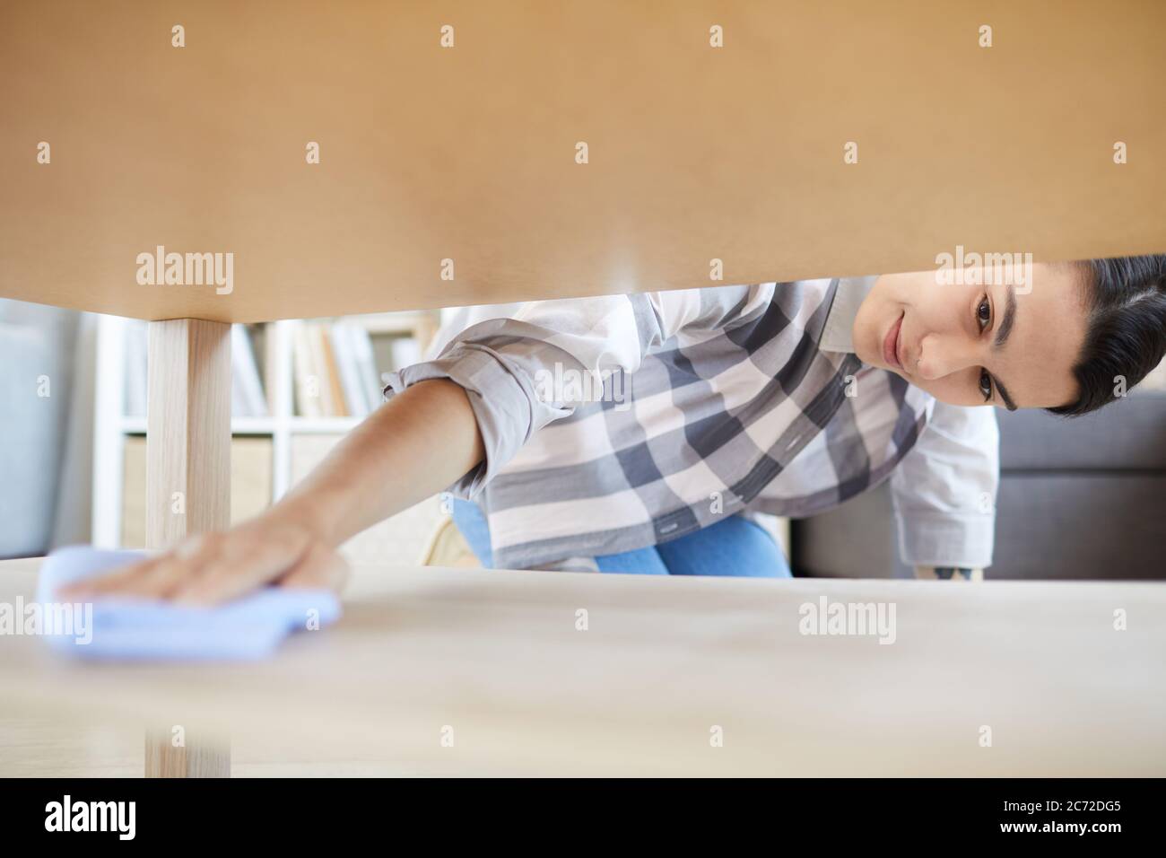 Young housewife using rag to wipe the dust under the table in the room ...