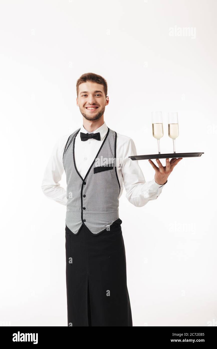 Young cheerful waiter in uniform happily looking in camera holding tray ...