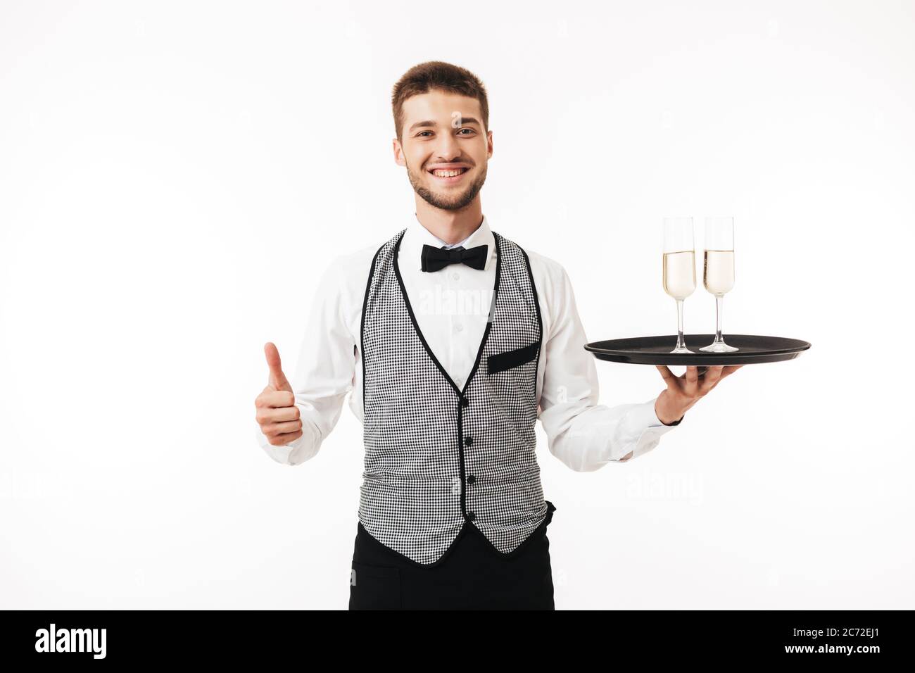 Young smiling waiter in uniform happily looking in camera holding tray ...
