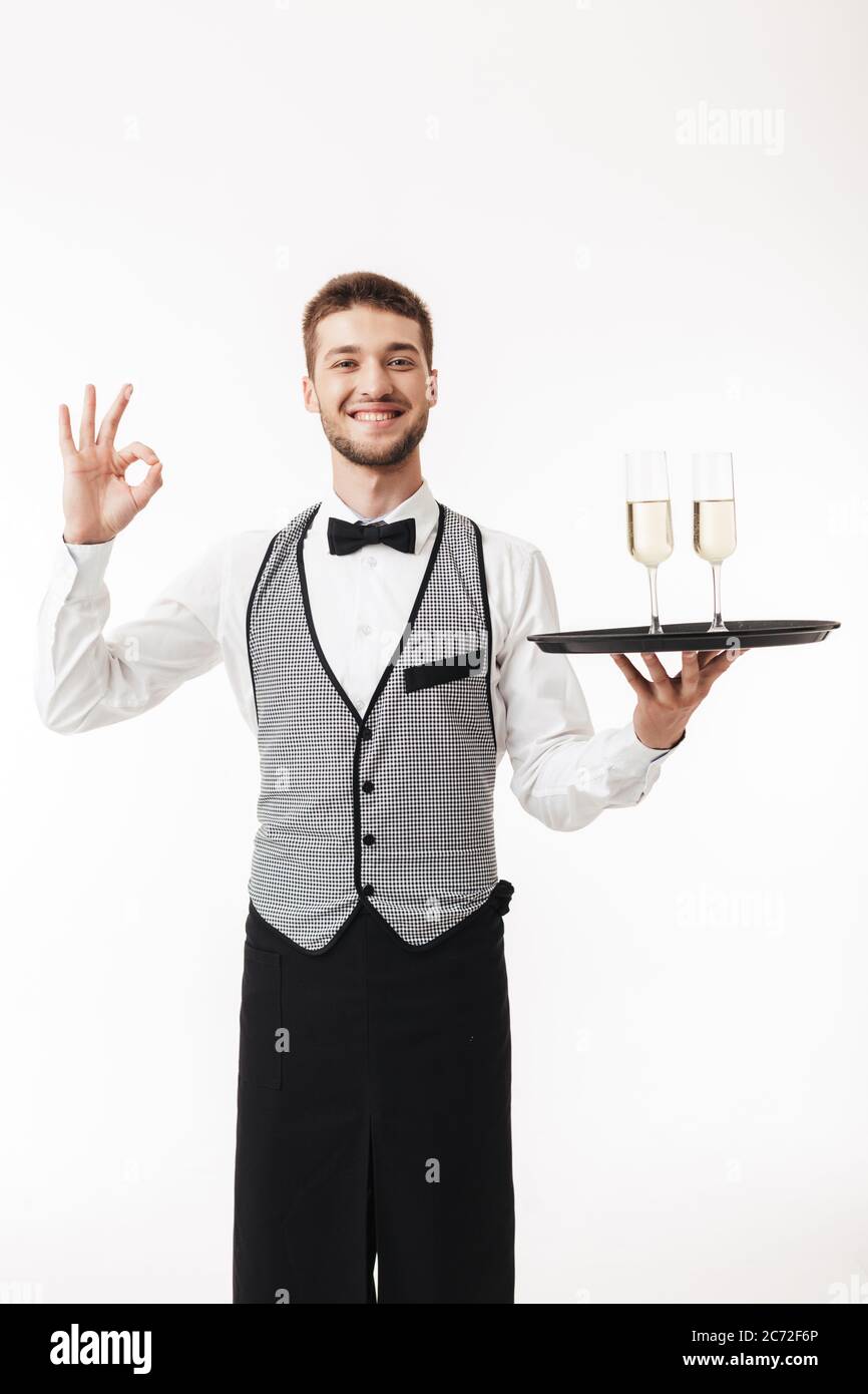 Young smiling waiter in uniform joyfully looking in camera holding tray ...