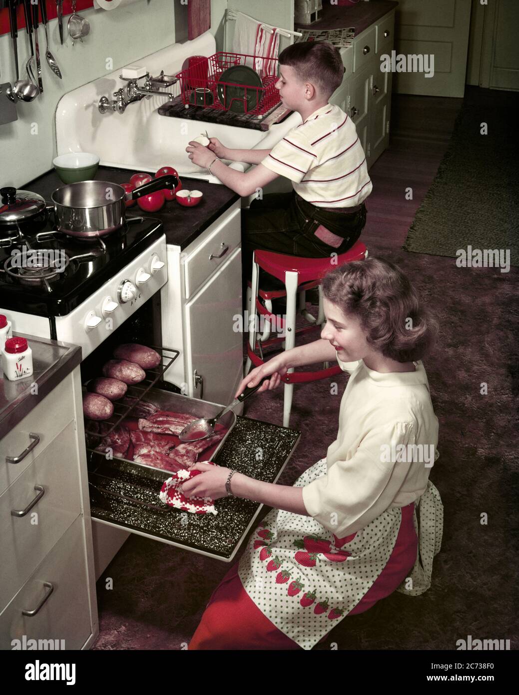 1950s KITCHEN SMILING TEENAGE GIRL SISTER KNEELING BY OVEN ROASTING ...