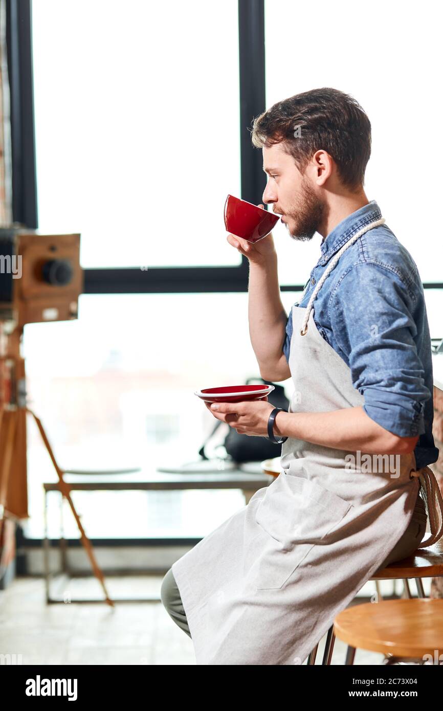 Beautiful calm barman wears white neat apron, drinks tasty aromatic ...