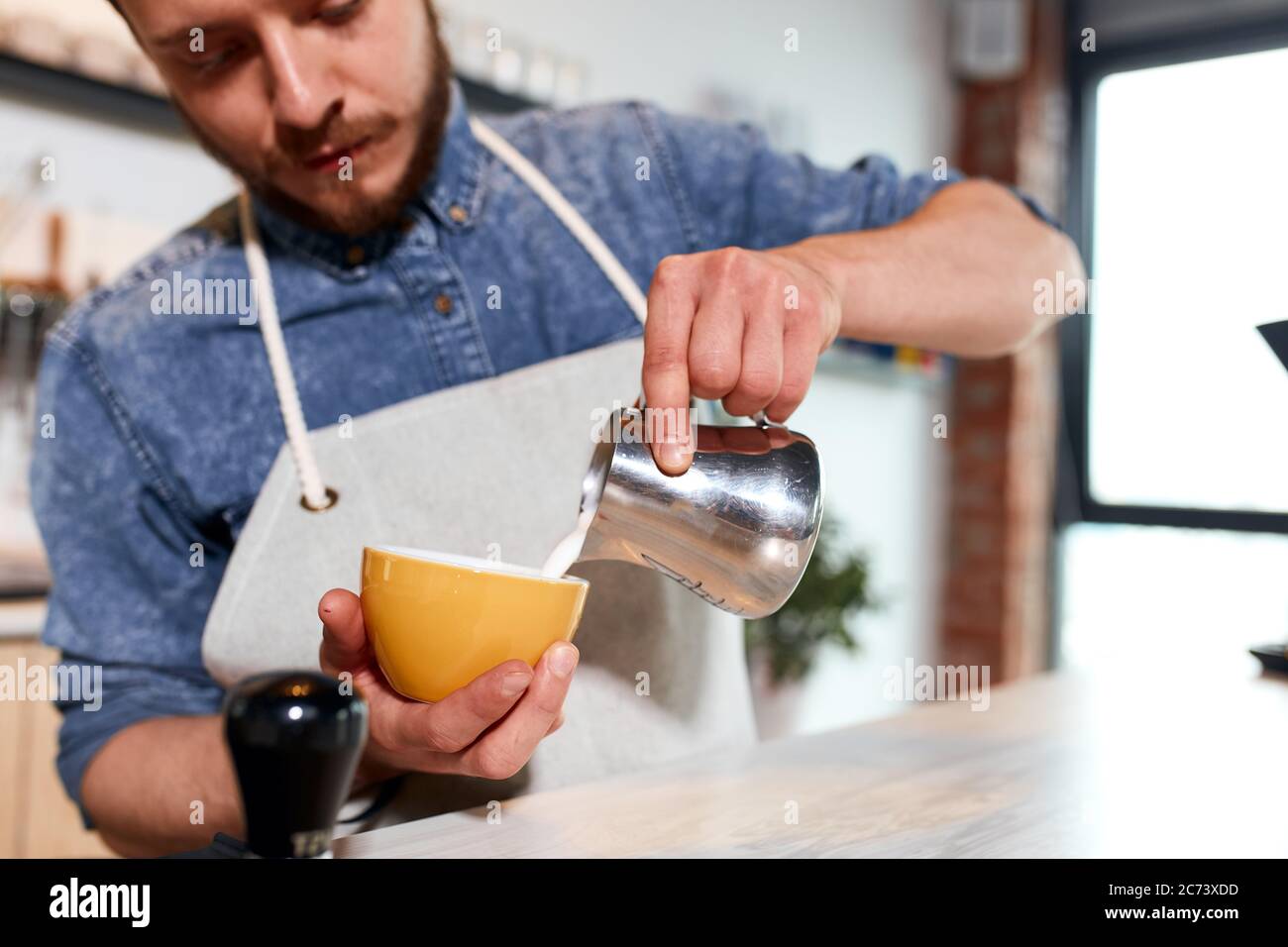 Confident concentrated bartender in neat apron pours steamed hot milk ...