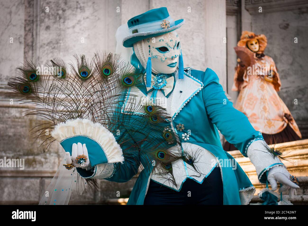A blue mask during Carnival in Venice waves a fan with peacock feathers ...
