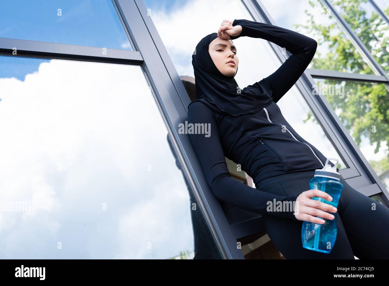 low angle view of tired muslim woman in hijab holding sports bottle ...