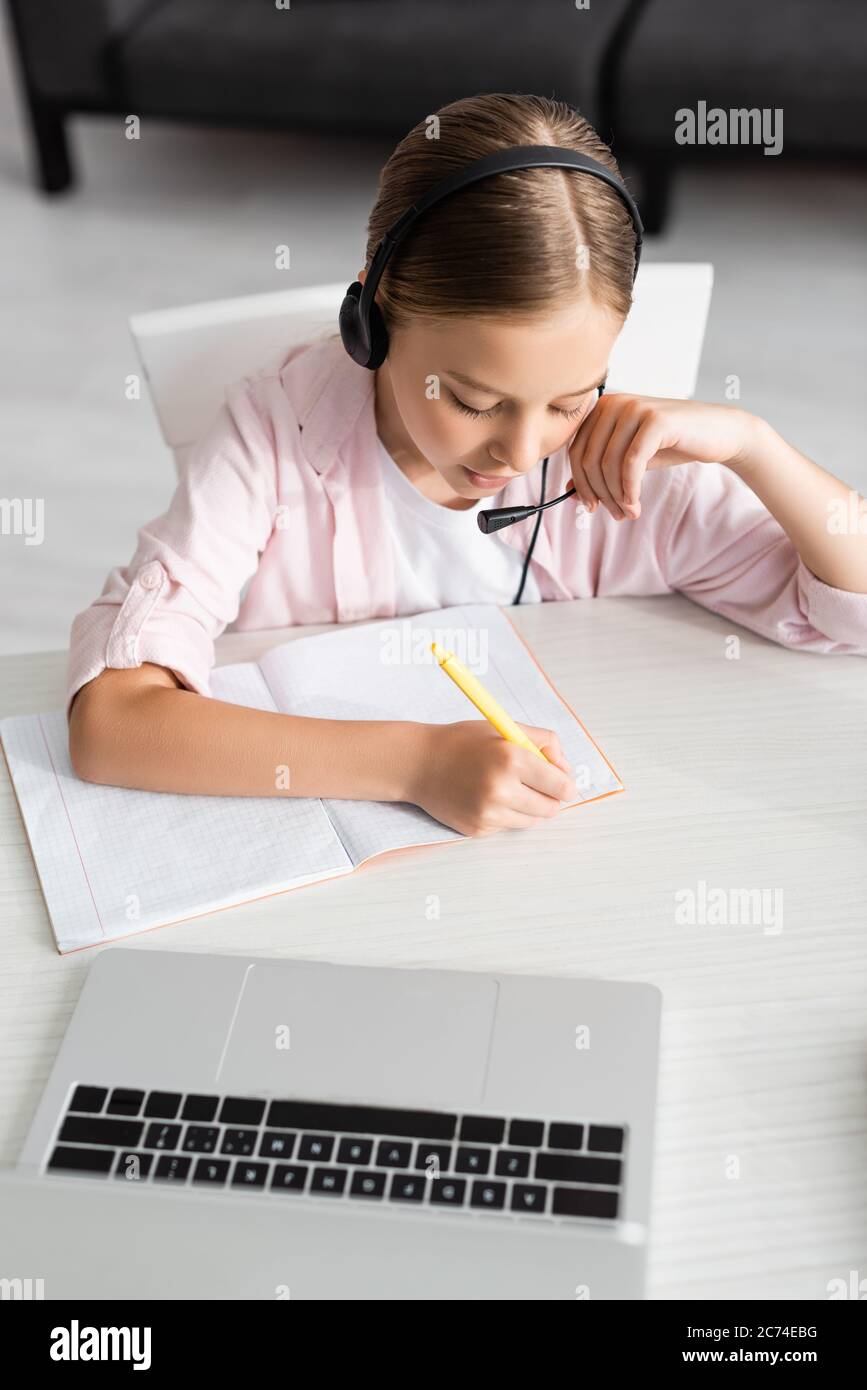 Overhead view of kid in headset writing on notebook near laptop on ...