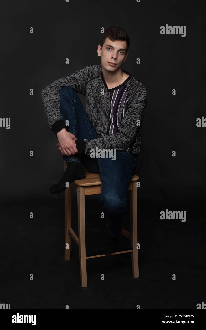 Portrait of serious young man is sitting on chair with one leg on it on ...