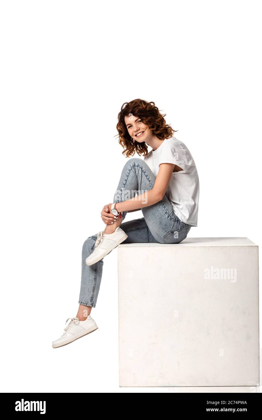 attractive woman in jeans sitting on cube and smiling isolated on white ...