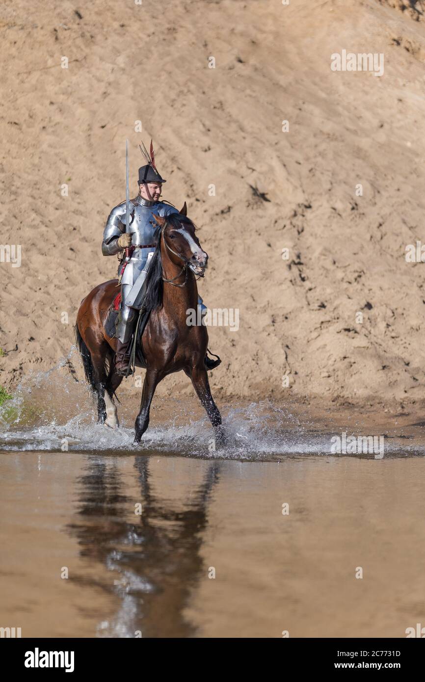 A young adult man in knightly armor rides a horse on a river along a ...