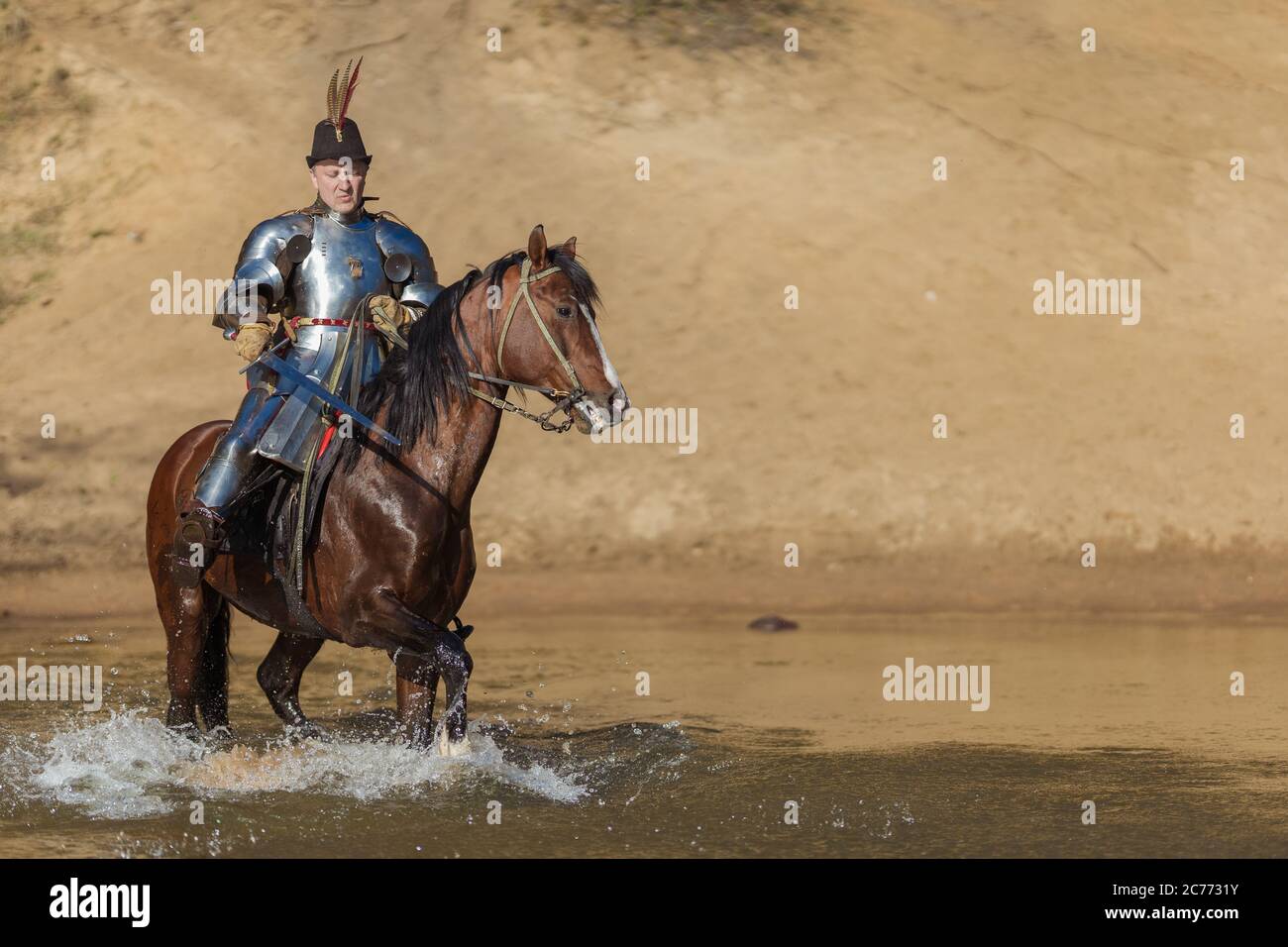 A young adult man in knightly armor rides a horse on a river along a ...