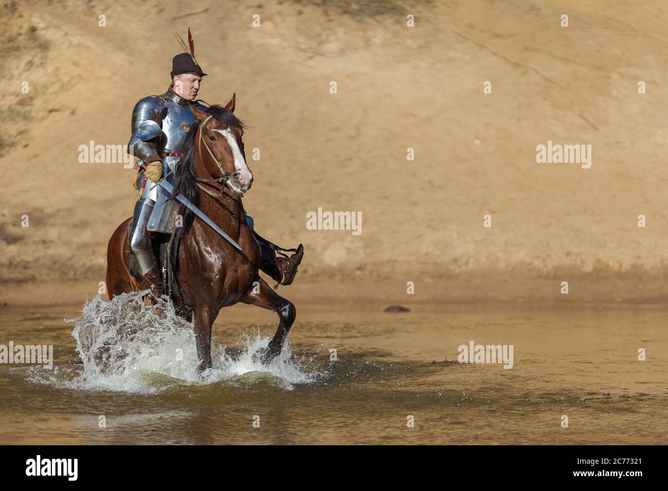 A young adult man in knightly armor rides a horse on a river along a ...