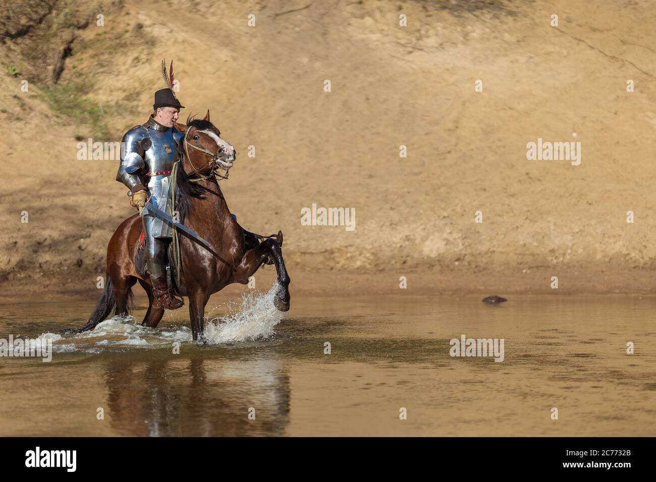 A young adult man in knightly armor rides a horse on a river along a ...