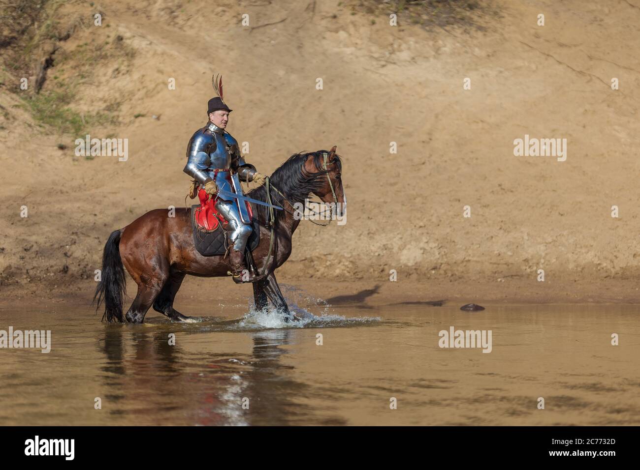 A young adult man in knightly armor rides a horse on a river along a ...
