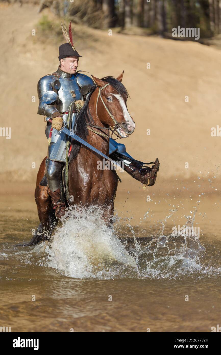 A young adult man in knightly armor rides a horse on a river along a ...