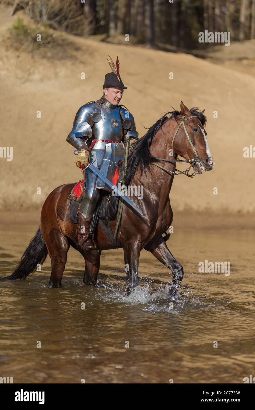 A young adult man in knightly armor rides a horse on a river along a ...