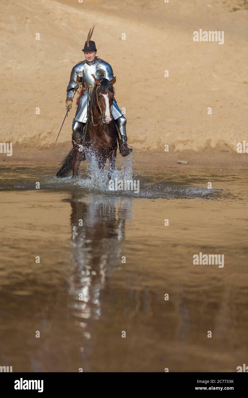 A young adult man in knightly armor rides a horse on a river along a ...