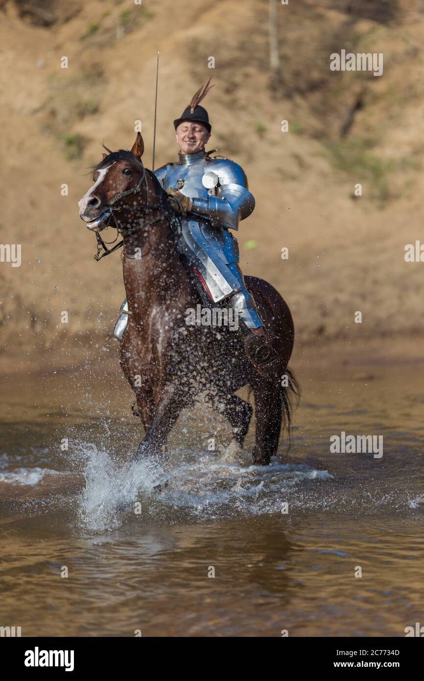 A young adult man in knightly armor rides a horse on a river along a ...