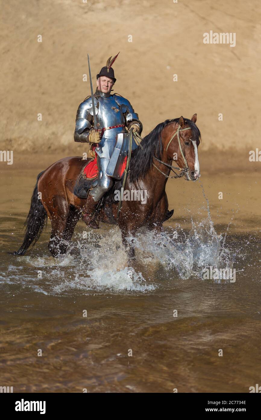 Adult man in ancient knight armor with a sword rides a horse on a river ...