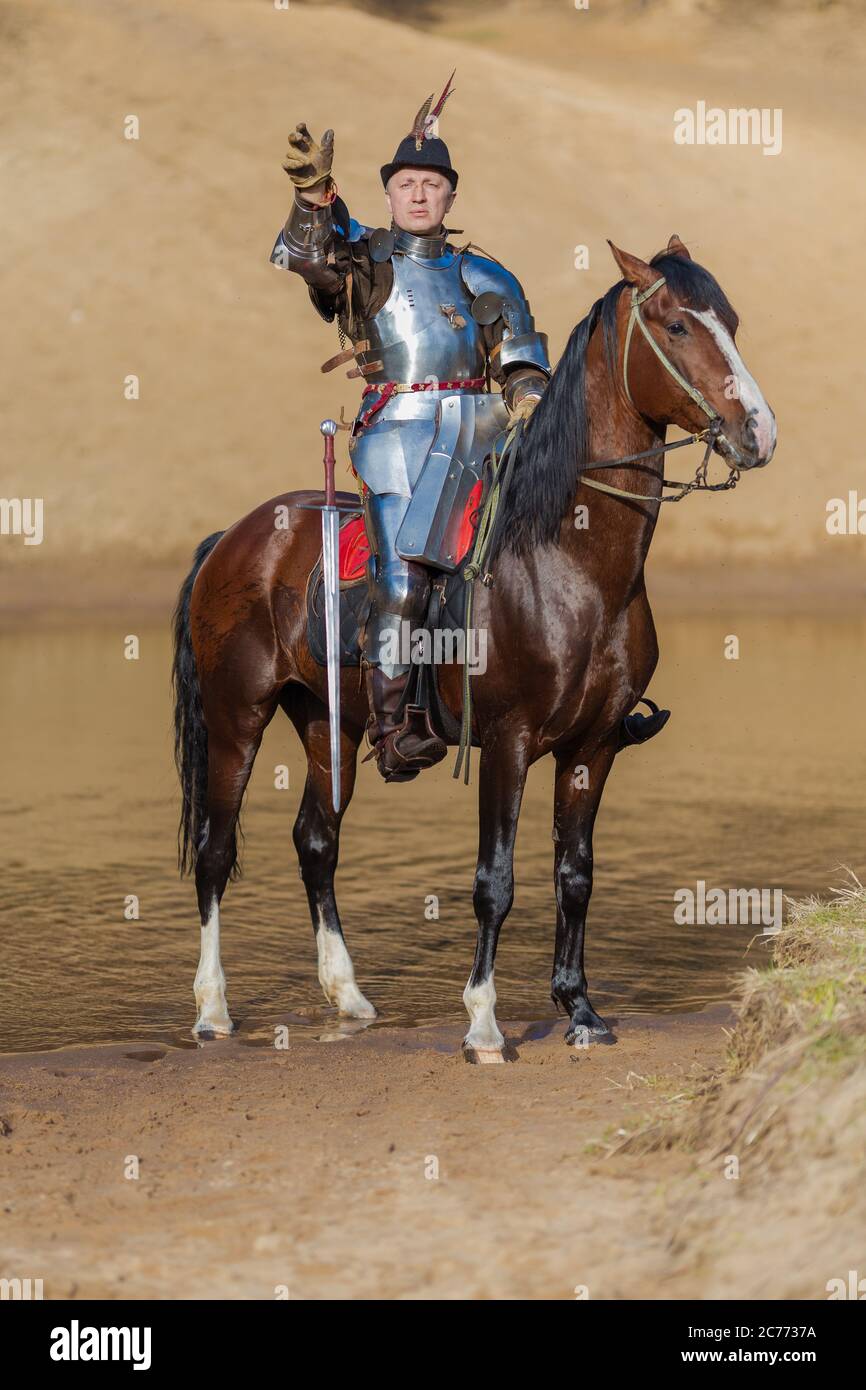 Adult man in ancient knight armor with a sword rides a horse on a river ...