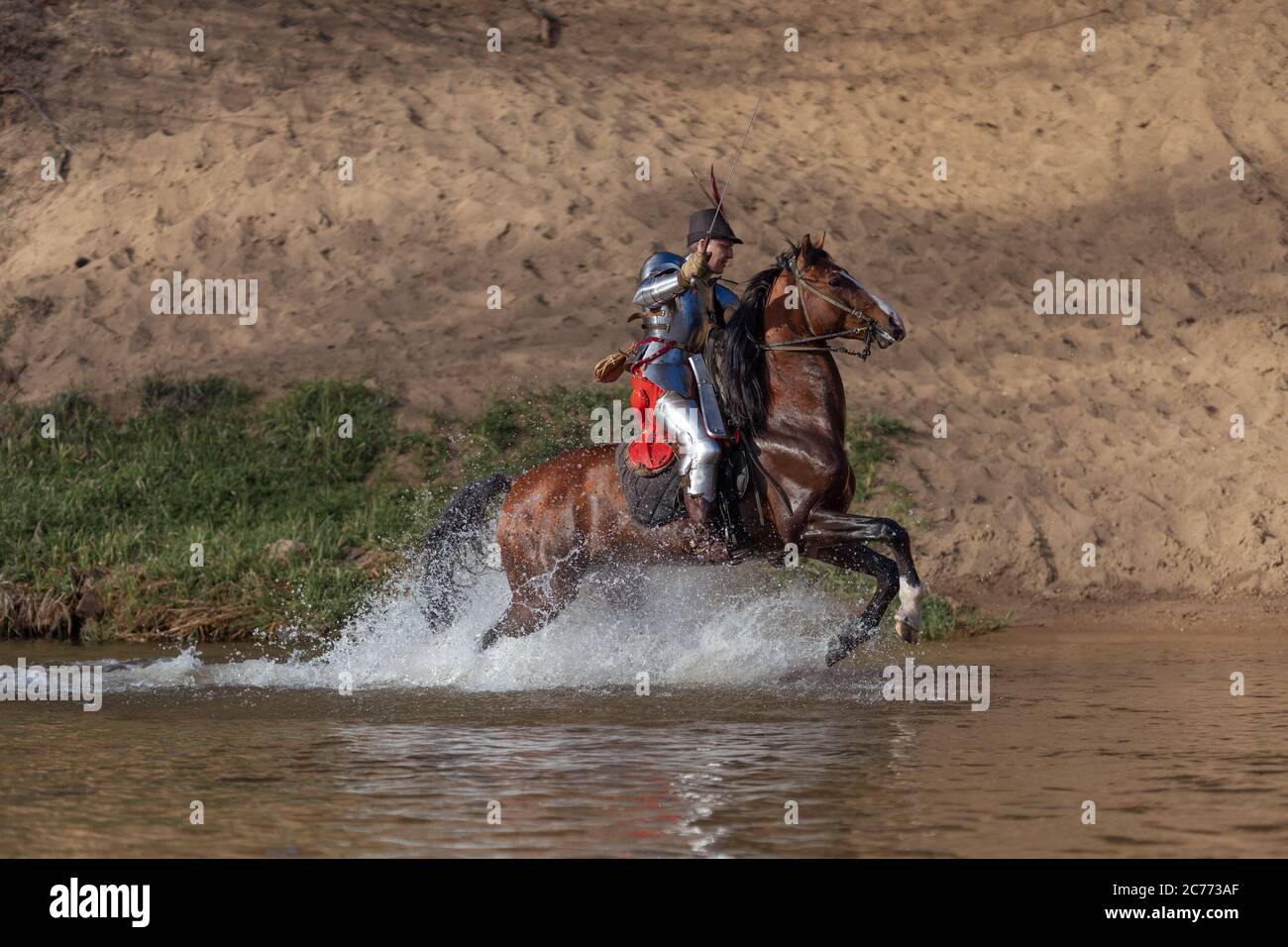 A young adult man in knightly armor rides a horse on a river along a ...