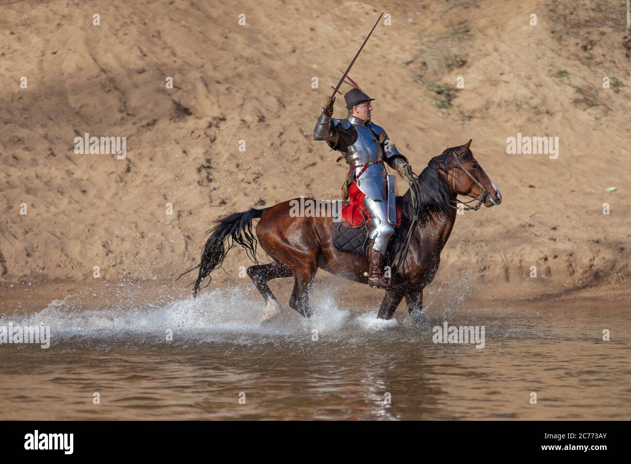 A young adult man in knightly armor rides a horse on a river along a ...