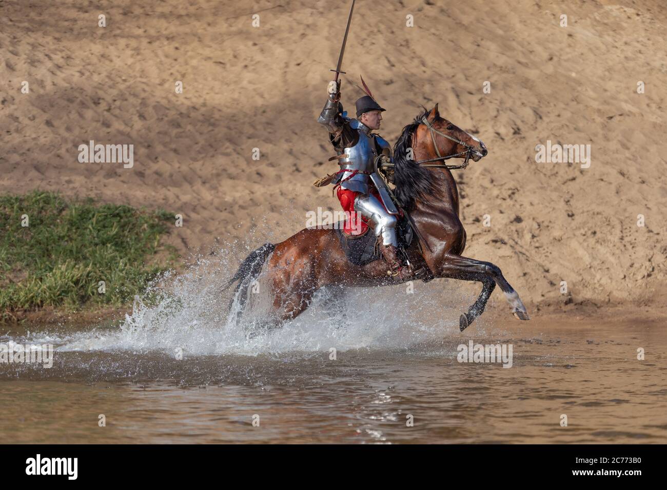 A young adult man in knightly armor rides a horse on a river along a ...
