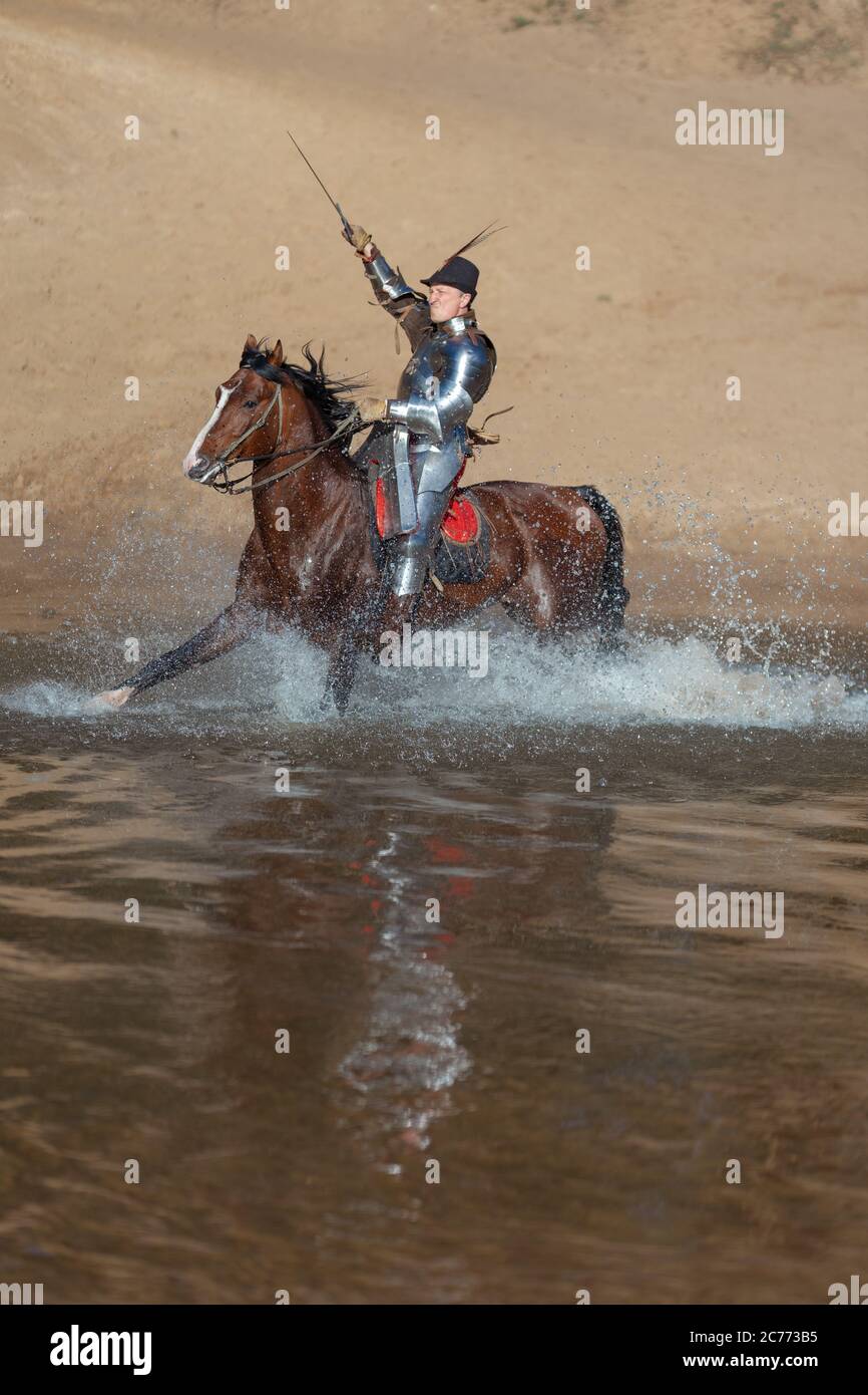 A young adult man in knightly armor rides a horse on a river along a ...