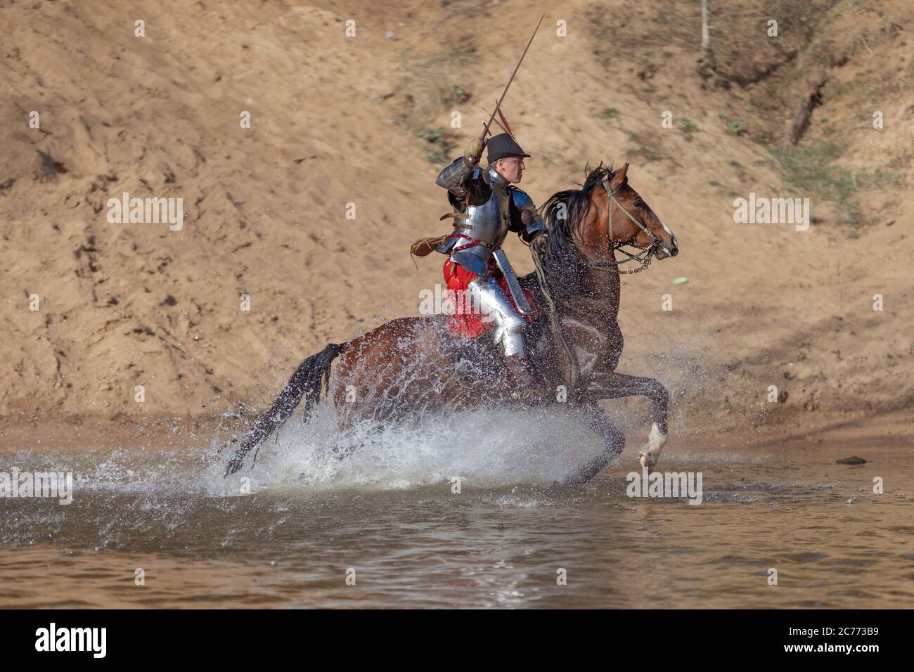 A young adult man in knightly armor rides a horse on a river along a ...