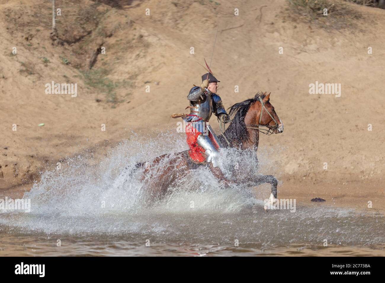 A young adult man in knightly armor rides a horse on a river along a ...