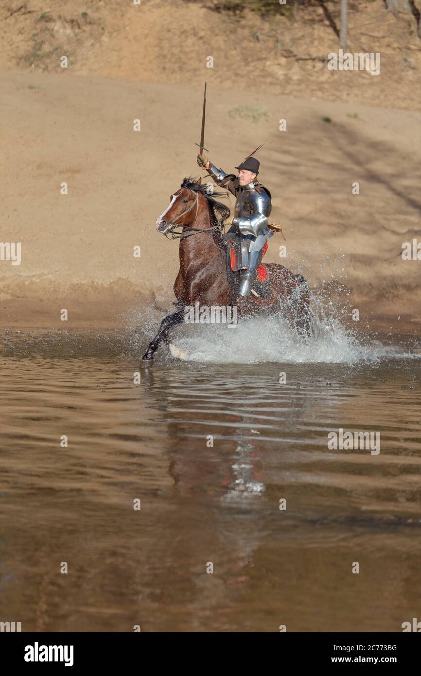 A young adult man in knightly armor rides a horse on a river along a ...