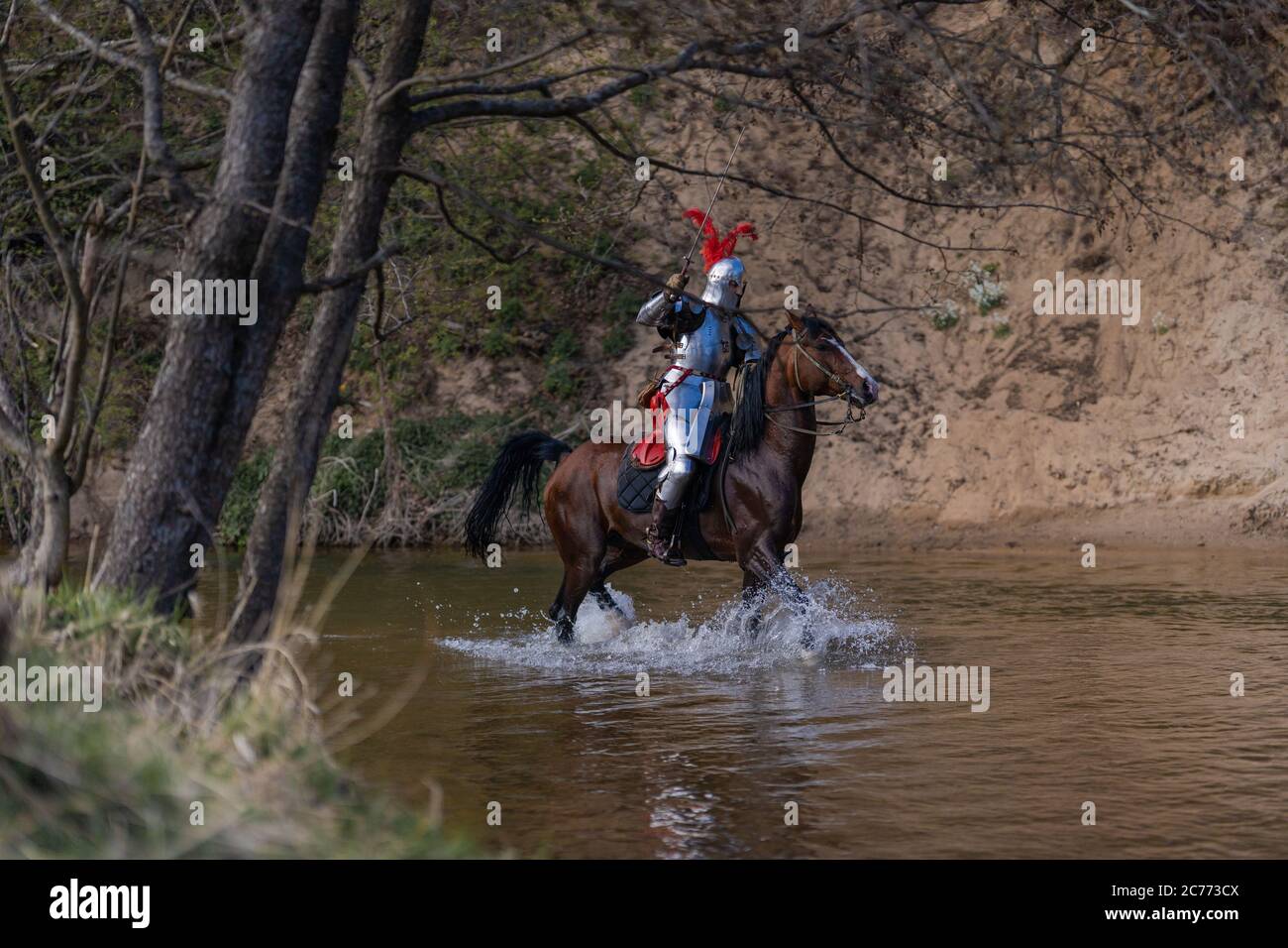 A young adult man in knightly armor rides a horse on a river along a ...