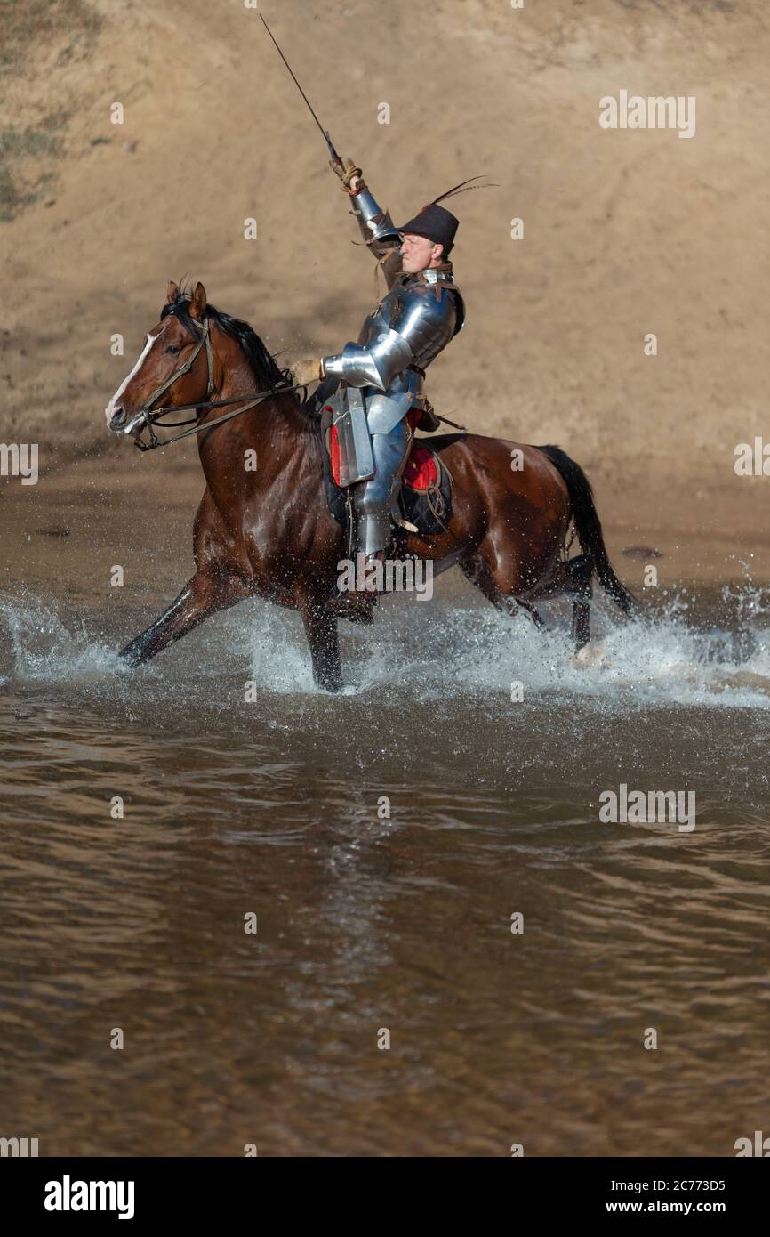 A young adult man in knightly armor rides a horse on a river along a ...